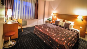 Quarto de hotel com uma cama grande, uma escrivaninha com cadeira, um banquinho pequeno, uma mesa de cabeceira com abajur e uma janela com cortinas.
