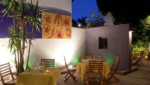 Uma área de refeições ao ar livre com mesas e cadeiras, cercada por plantas e uma parede com um mural floral.