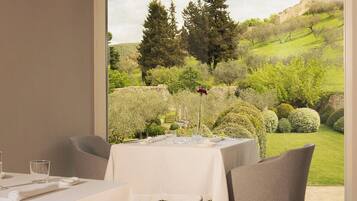 Uma área de jantar com uma mesa posta para dois, e uma janela com vista para um jardim paisagístico.