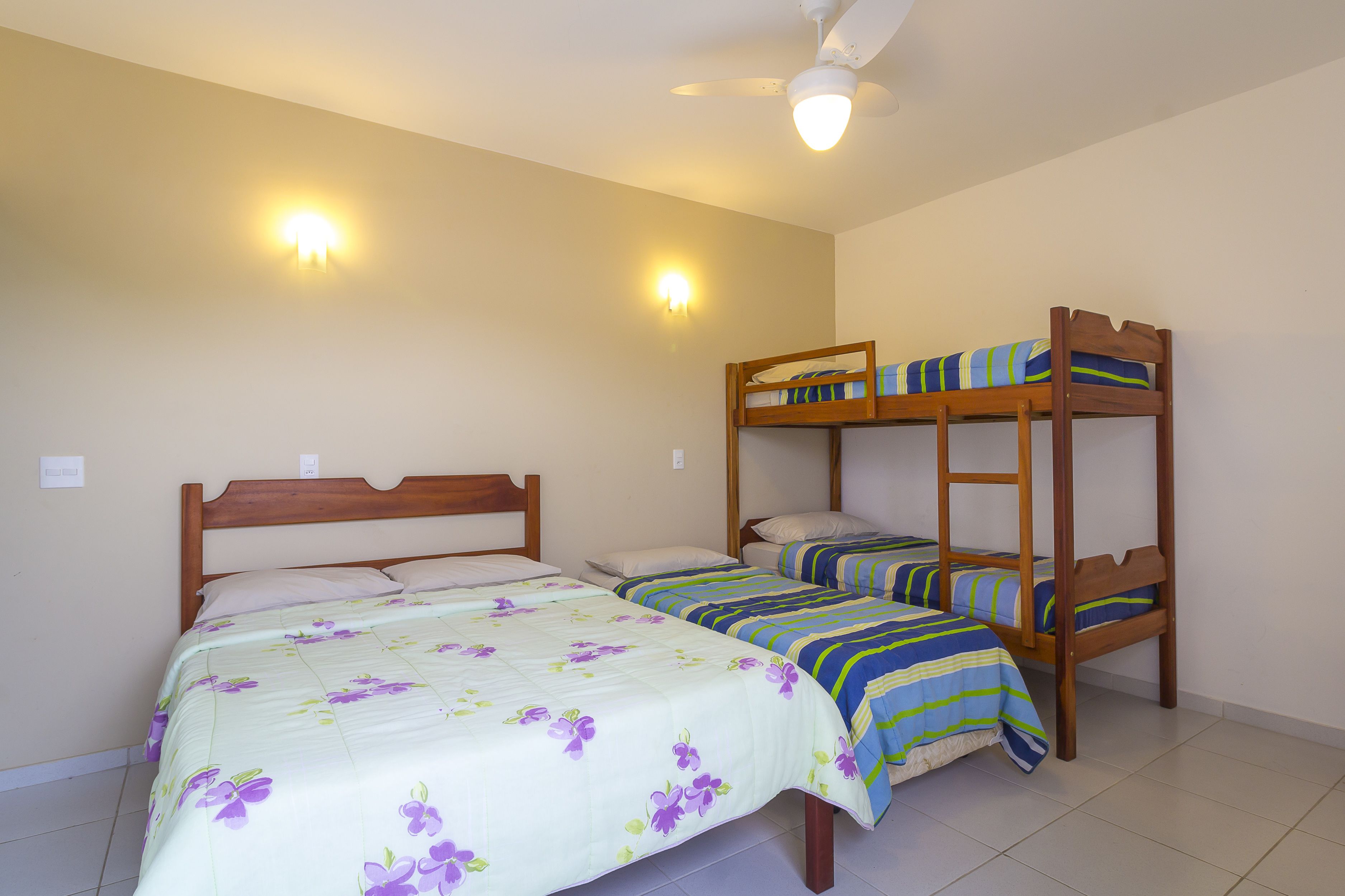 Quarto com uma cama de solteiro e duas camas beliche, um ventilador de teto e duas luminárias fixadas na parede.