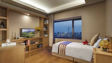Quarto de hotel moderno com uma cama grande, uma TV, uma mesa com uma xícara e papéis, e vista para a cidade através de uma janela ampla.