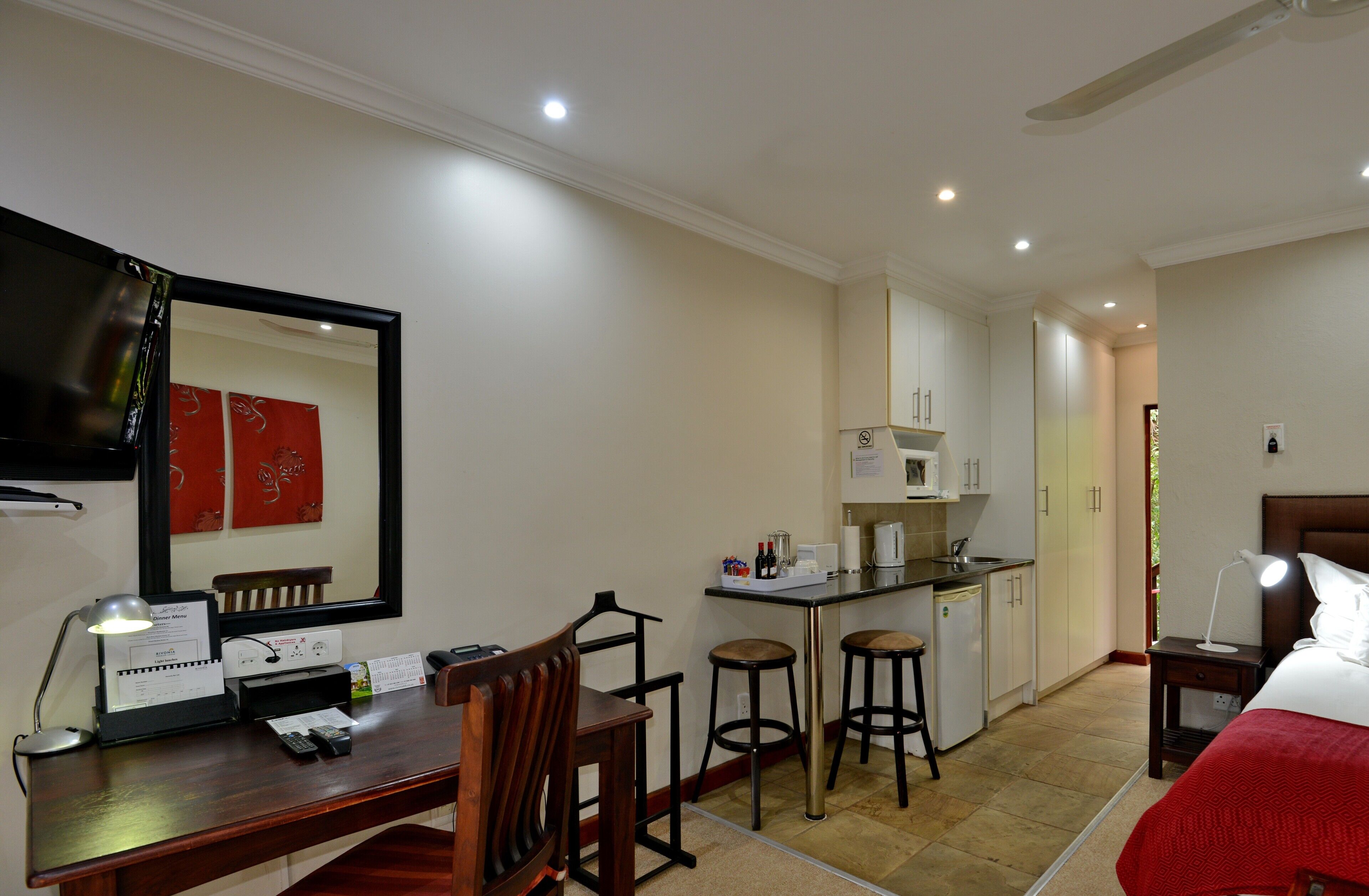 Quarto de hotel com escrivaninha, cadeira, dois bancos, cama, mesa de cabeceira, abajur, televisão e cozinha compacta.