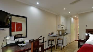 Quarto de hotel com escrivaninha, cadeira, dois bancos, cama, mesa de cabeceira, abajur, televisão e cozinha compacta.