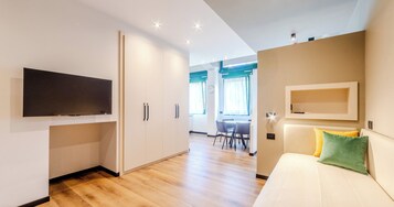 Quarto moderno com cama, televisão e área de refeições ao fundo.