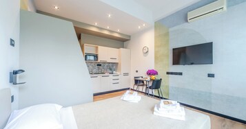 Quarto de hotel moderno com cama, mesa de jantar com cadeiras, área de cozinha e televisão na parede.