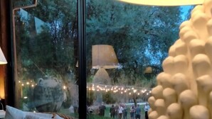 Um sofá de vime com almofadas brancas, uma luminária e vista para área externa com luzes de cordão e pessoas.
