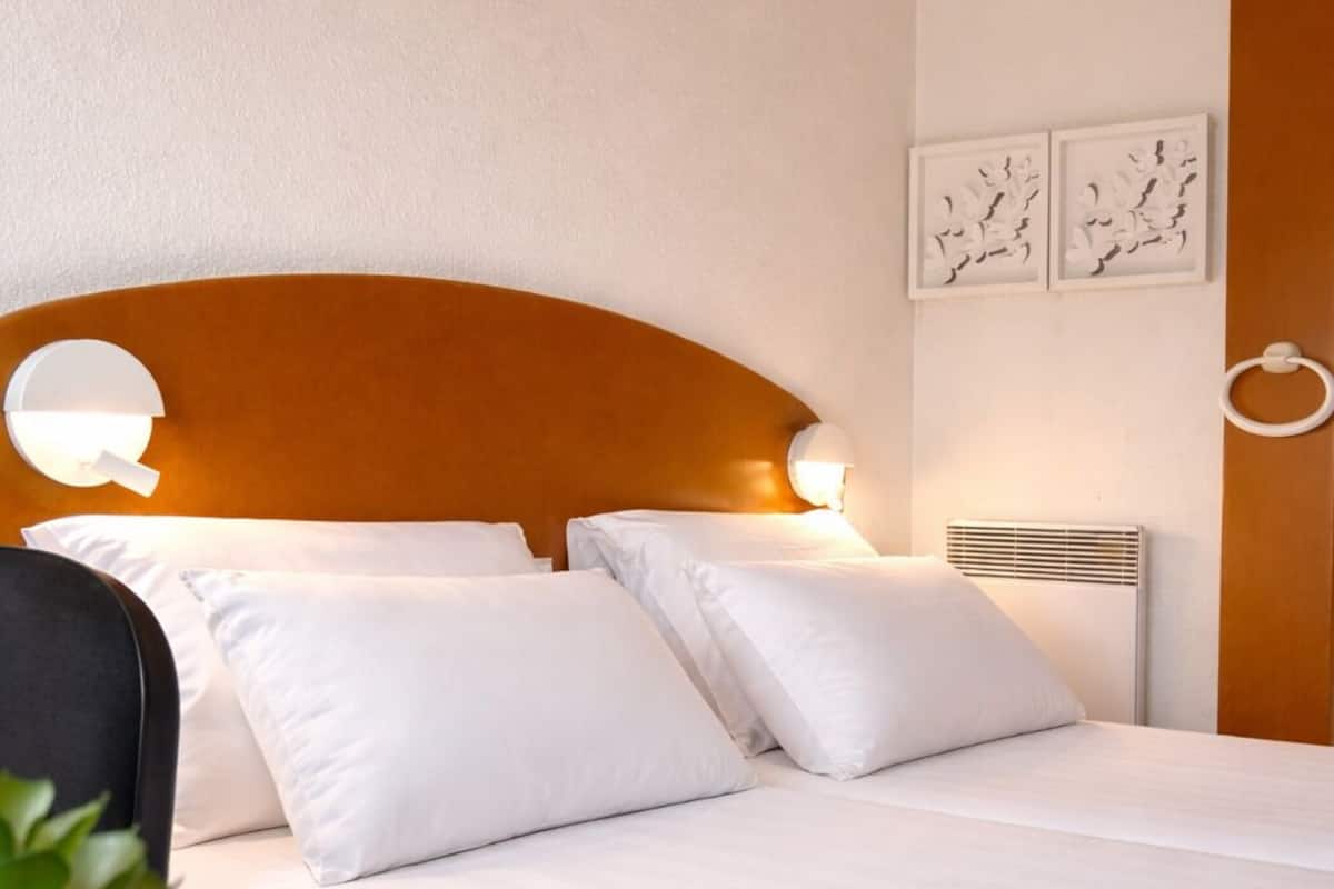 Quarto de hotel com cama, almofadas brancas, mesa de cabeceira vermelha e um vaso com planta.
