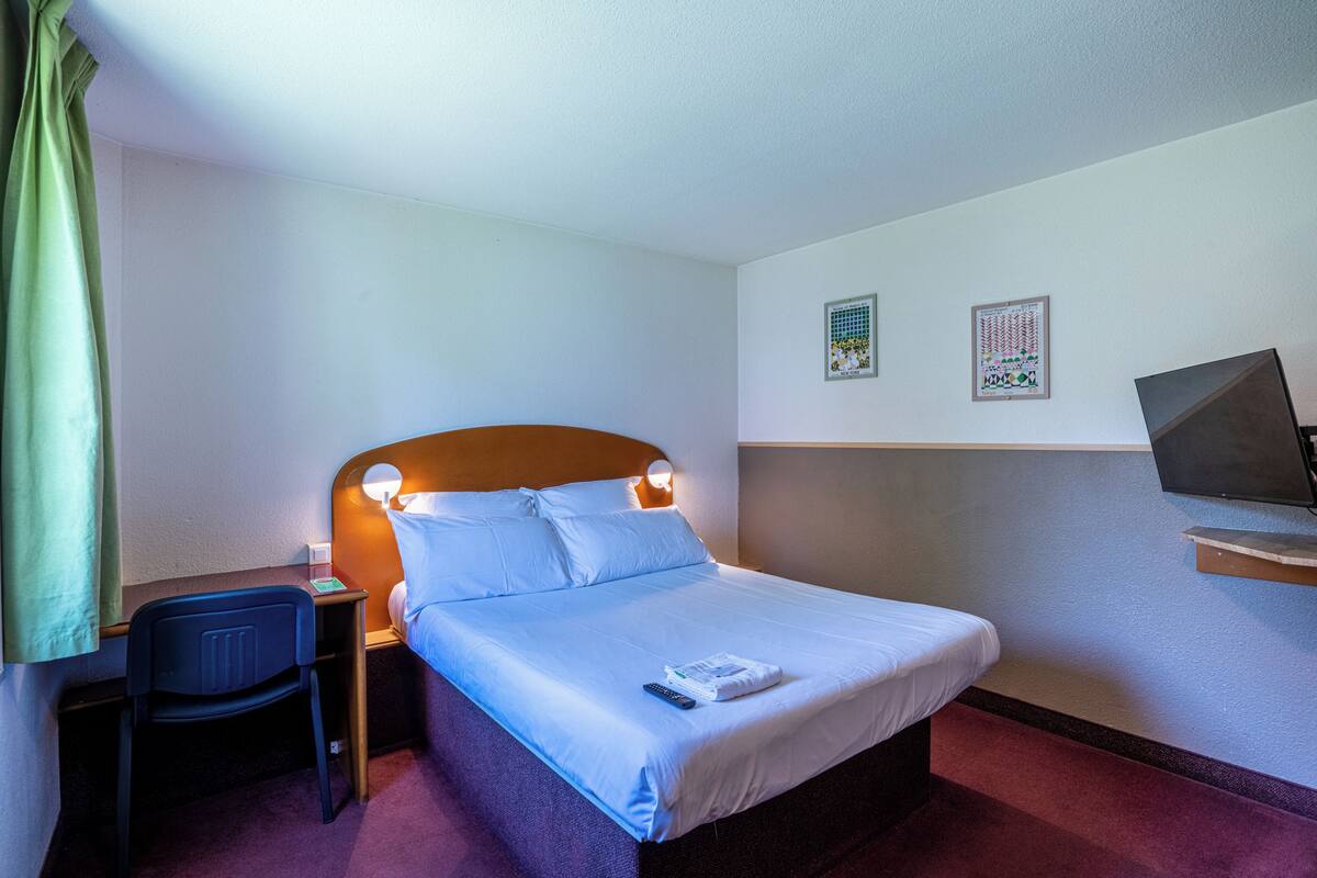Quarto de hotel com cama, escrivaninha, cadeira, televisão e duas decorações na parede.