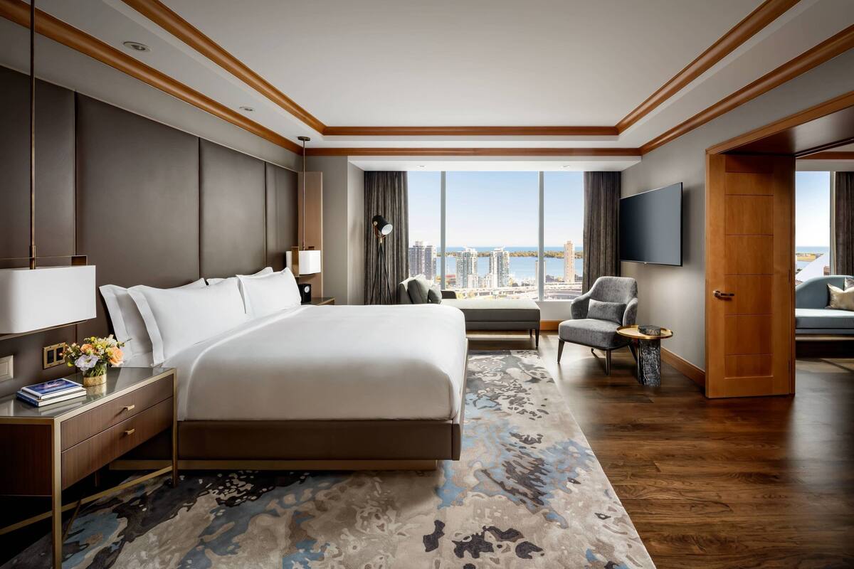 Quarto de hotel moderno com uma cama grande, área de estar, televisão e vista para a cidade.