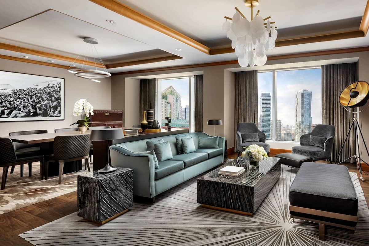 Uma sala de estar moderna com sofá, mesa de centro e cadeiras, grandes janelas com vista para a cidade, e um espaço de jantar com mesa e cadeiras.