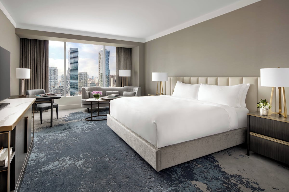 Quarto de hotel moderno com uma cama grande, uma escrivaninha, uma cadeira e vista para o horizonte da cidade.
