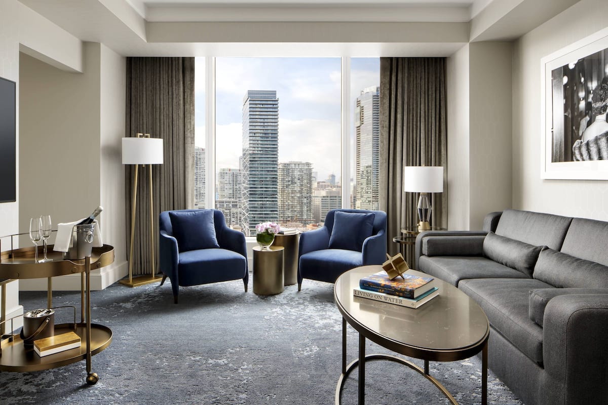 Quarto de hotel moderno com vista para a cidade, com um sofá cinzento, poltronas azuis, uma mesa de centro redonda e uma mesinha lateral dourada com um tabuleiro.
