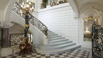 Uma escadaria imponente com grades de metal preto ricamente decoradas, piso xadrez e parede de mármore.