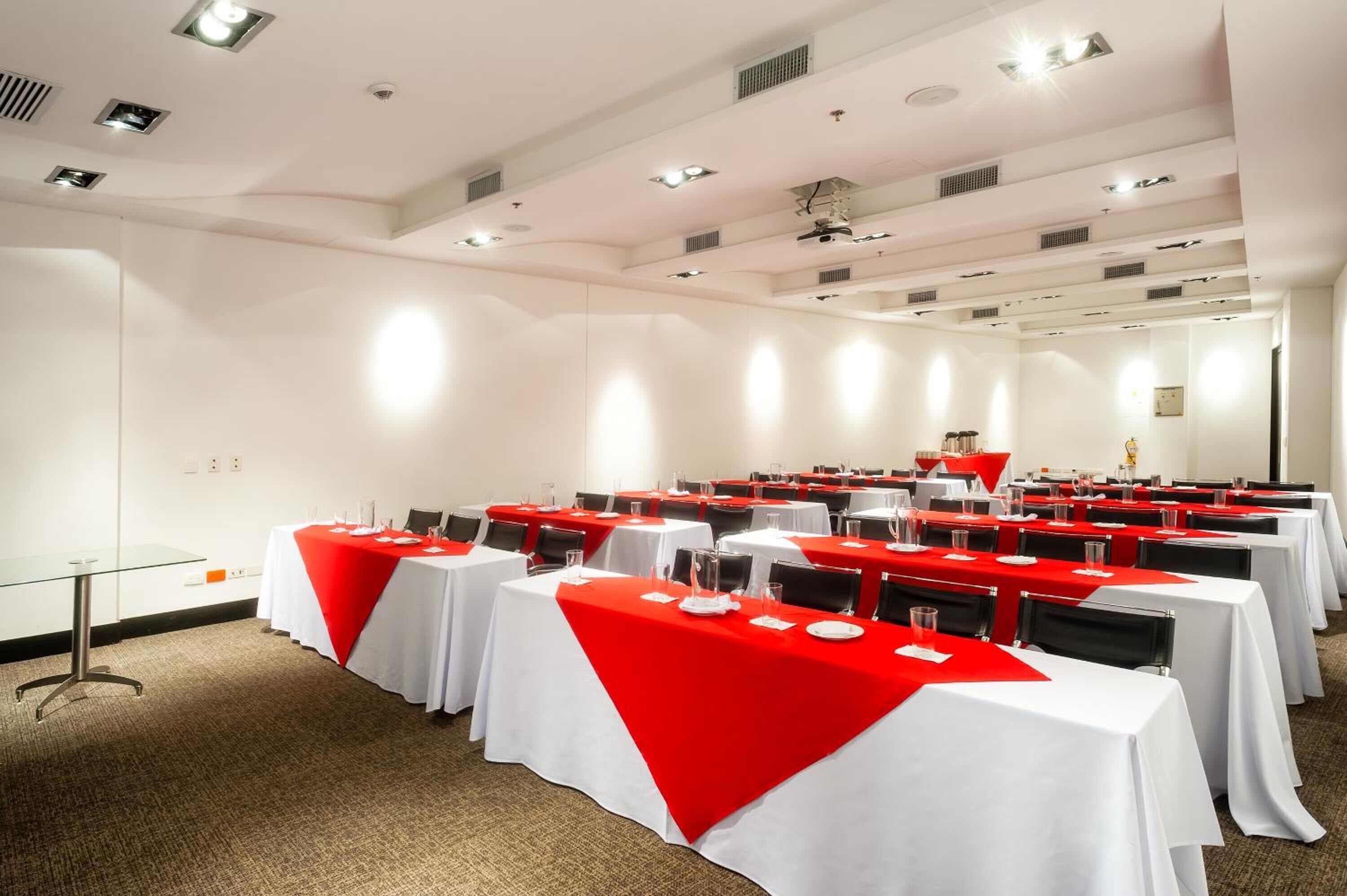 Sala de conferência com fileiras de mesas, cada uma coberta por toalhas brancas e jogos americanos vermelhos.