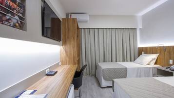 Quarto de hotel com escrivaninha de madeira, duas camas, uma televisão e um aparelho de ar condicionado.