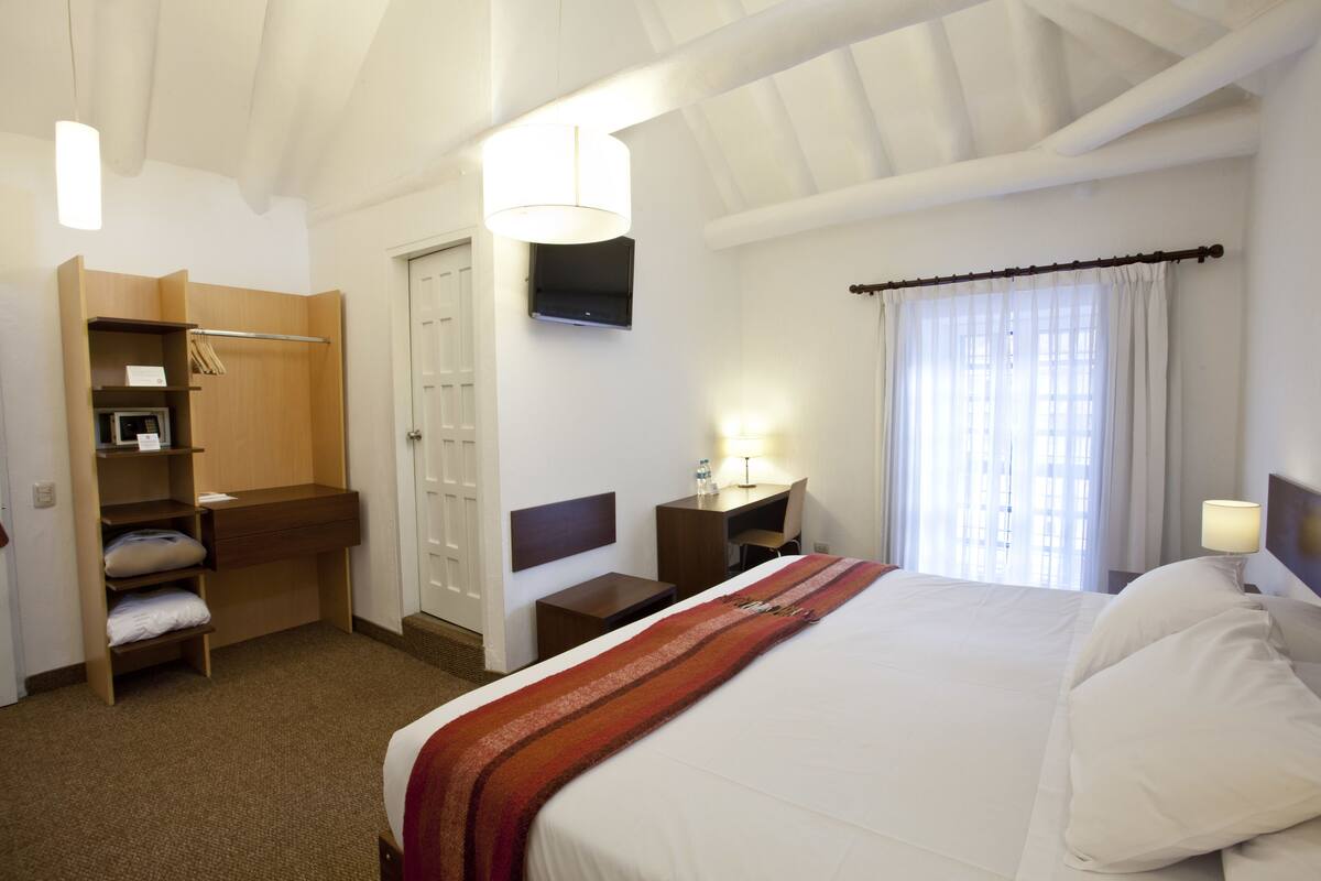 Quarto de hotel com cama, escrivaninha, cadeira, guarda-roupa e televisão.