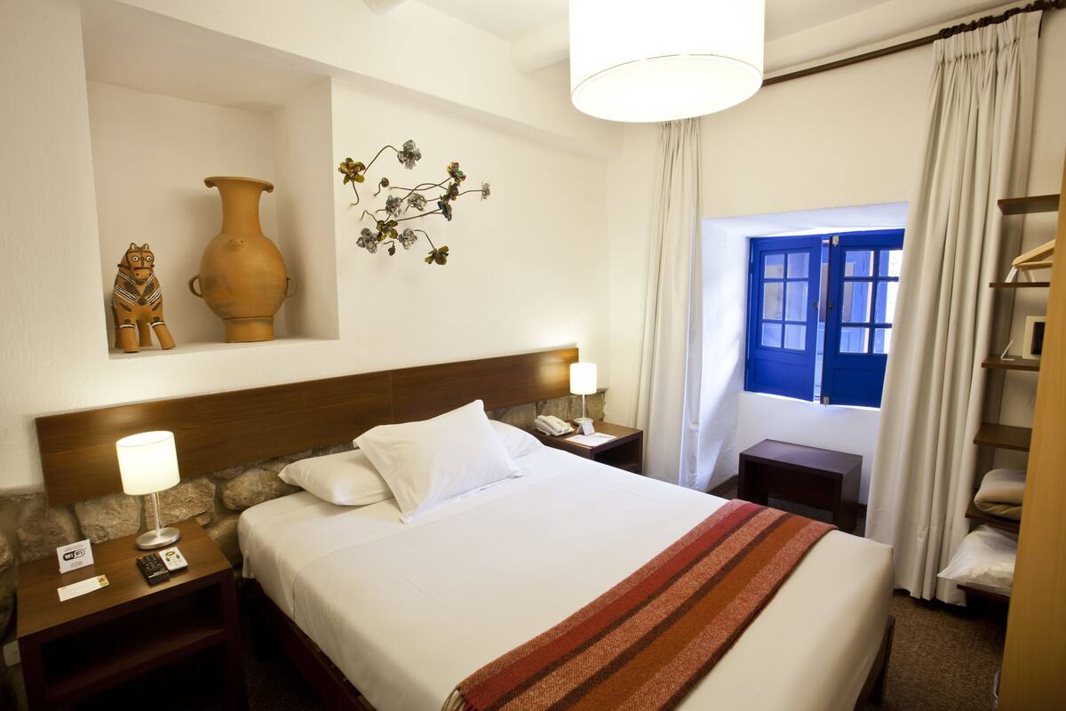 Quarto de hotel com cama, mesas de cabeceira, um vaso, decoração na parede e uma janela com cortinas.