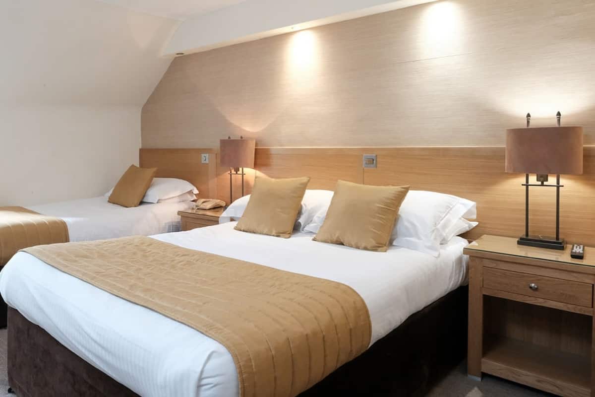 Quarto de hotel com duas camas, cabeceiras de madeira, luminárias de mesa e uma mesa de cabeceira com gaveta.