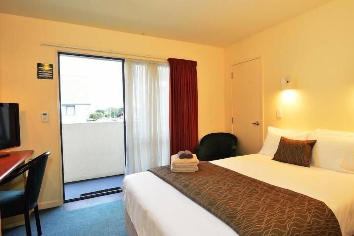Quarto de hotel com cama, escrivaninha, cadeira, televisão e porta de correr para a varanda.