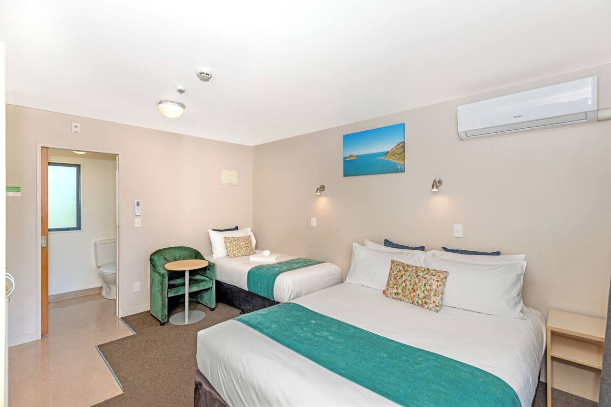 Quarto de hotel com duas camas, uma mesinha, uma cadeira, um quadro e um aparelho de ar condicionado.