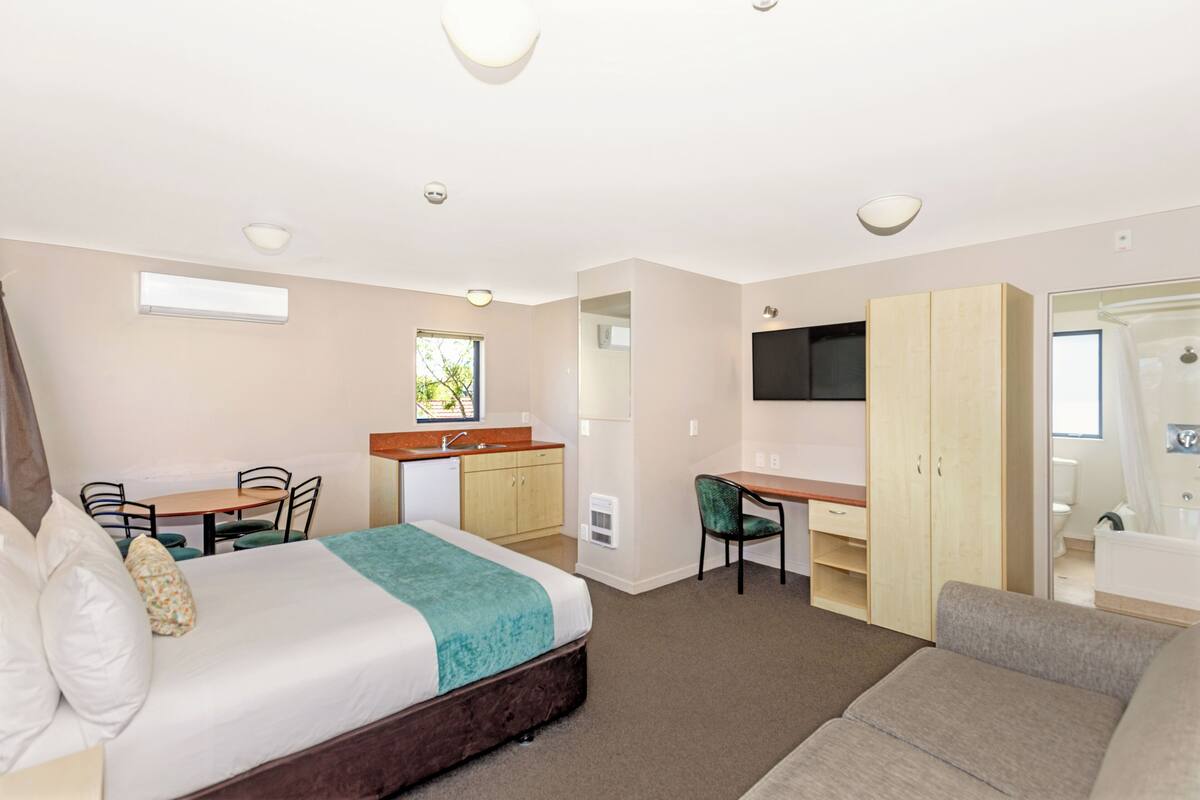 Quarto de hotel com cama, escrivaninha, cadeira, sofá, televisão, banheiro e janela.