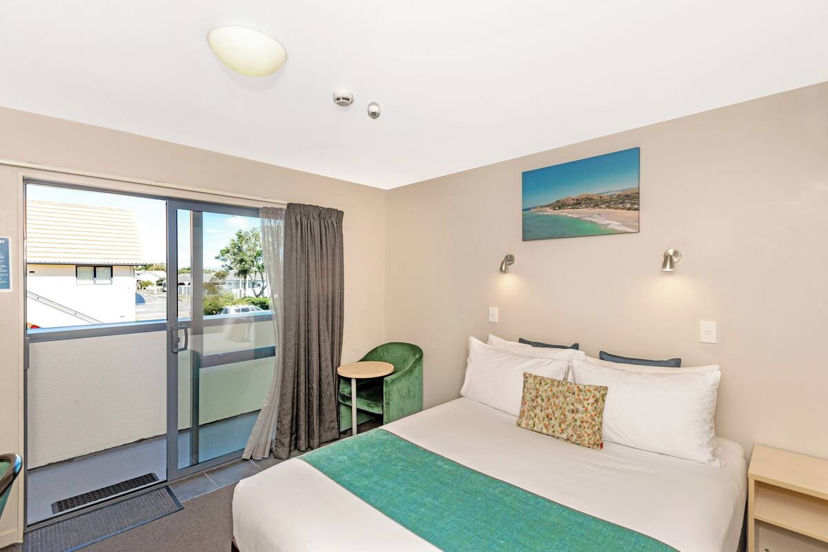 Quarto de hotel com cama, uma mesinha, uma cadeira, vista para o exterior e um quadro na parede.