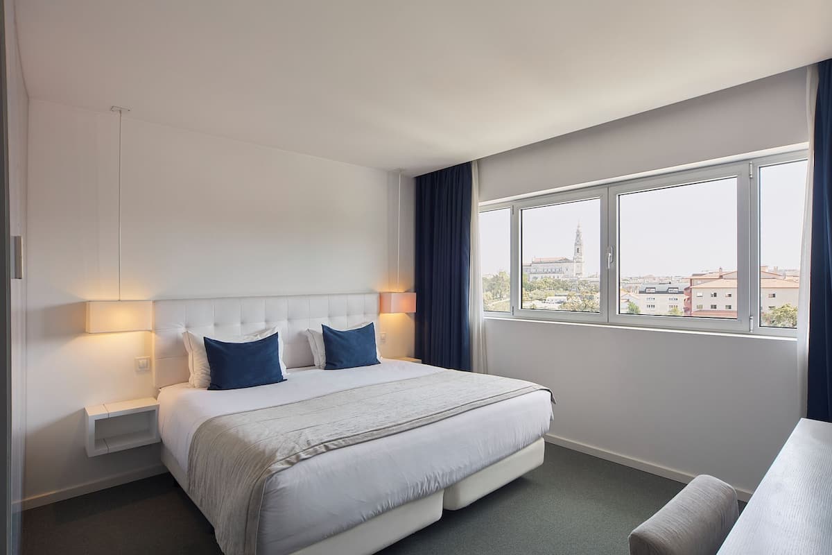 Quarto de hotel com uma cama grande, uma mesa de cabeceira, um abajur, uma janela com vista para a cidade e uma cadeira.