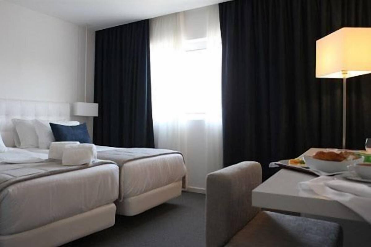 Quarto de hotel com cama, duas mesinhas de cabeceira, escrivaninha, cadeira e janela com cortinas.
