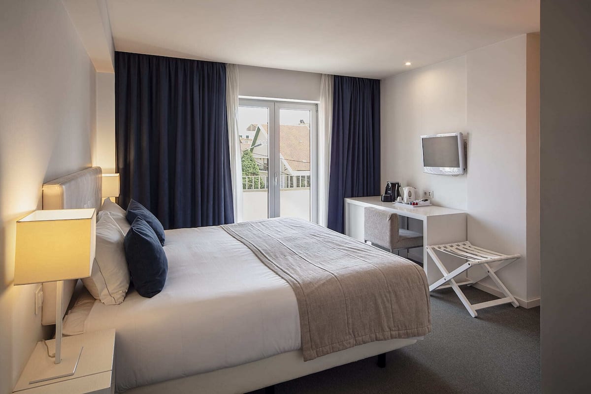 Quarto de hotel com cama, escrivaninha, cadeira, televisão e cortinas.