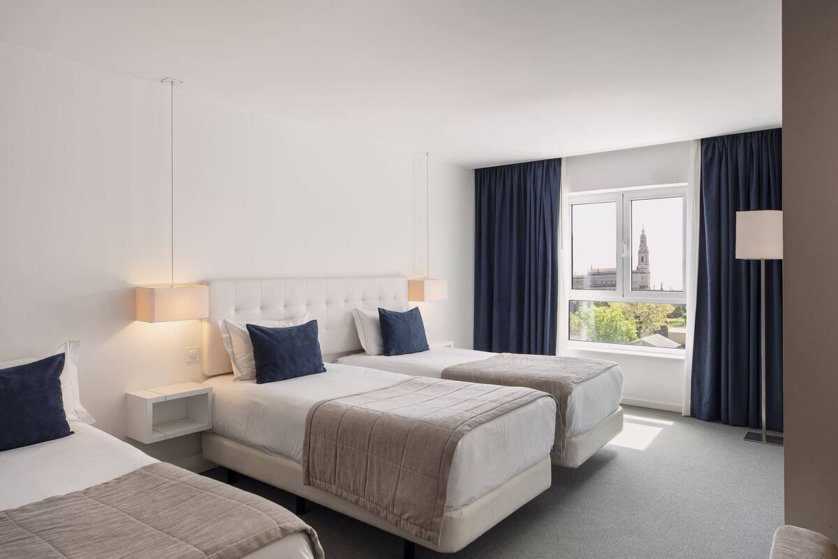 Quarto de hotel com duas camas, janela com cortinas, luminária e vista para um edifício.