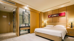 Quarto de hotel moderno com uma cama grande, banheiro com espelho e pia, e um quadro abstrato vermelho na parede.