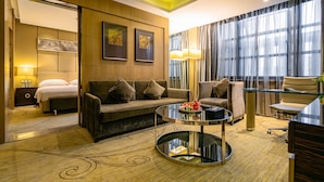Quarto de hotel moderno com sofá, mesa de centro de vidro, escrivaninha e uma janela grande com cortinas.