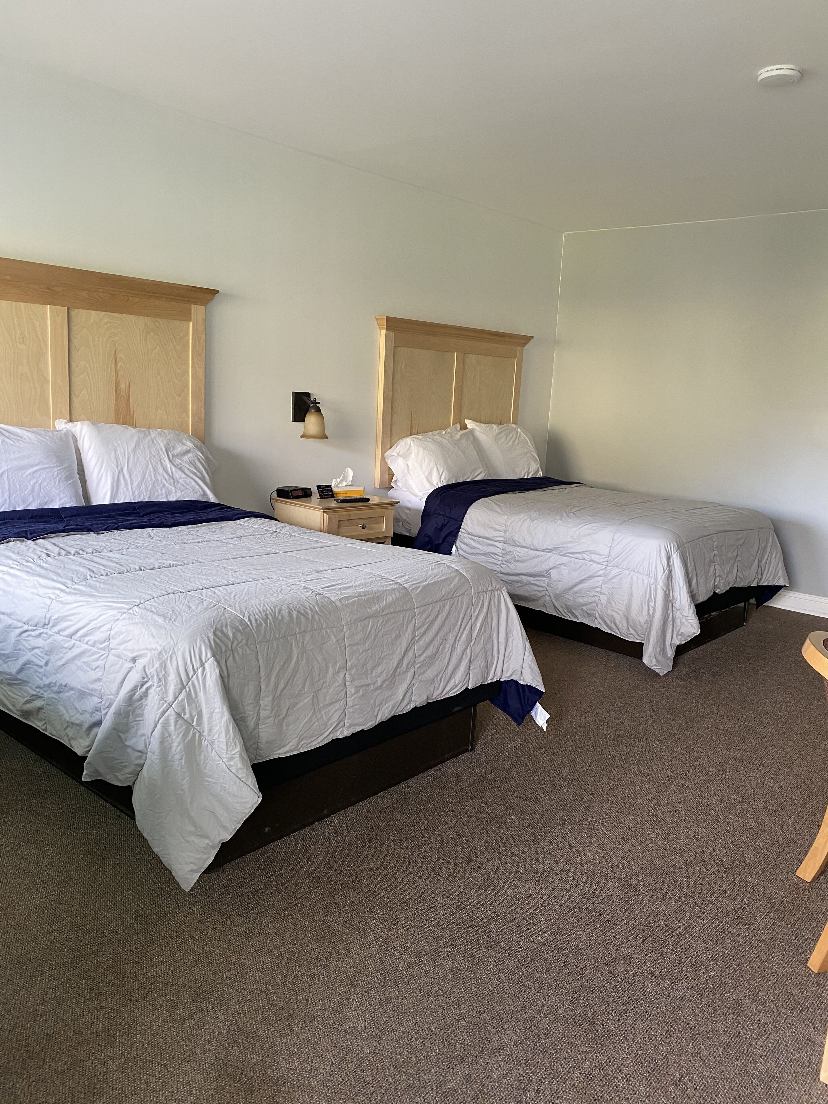 Quarto de hotel com duas camas, cada uma com cabeceira de madeira, uma mesa de cabeceira com abajur e piso coberto por carpete.