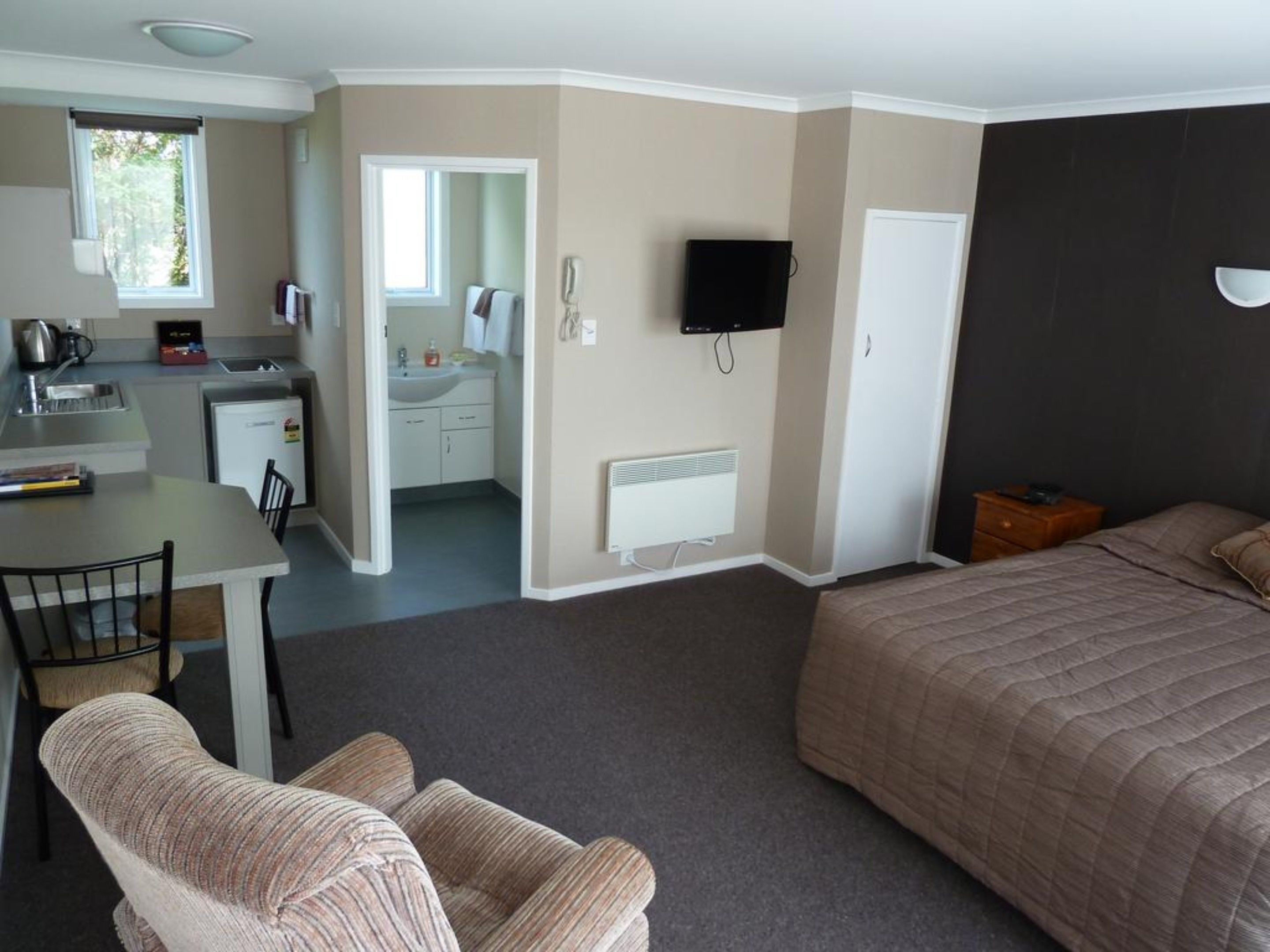 Quarto de hotel compacto com uma cama, escrivaninha, cadeira, cozinha compacta e televisão presa à parede.