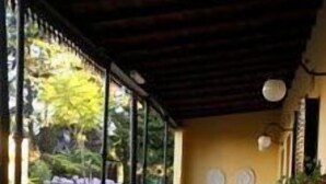 Uma área externa coberta com piso de azulejos, cadeiras de madeira e uma mesa.