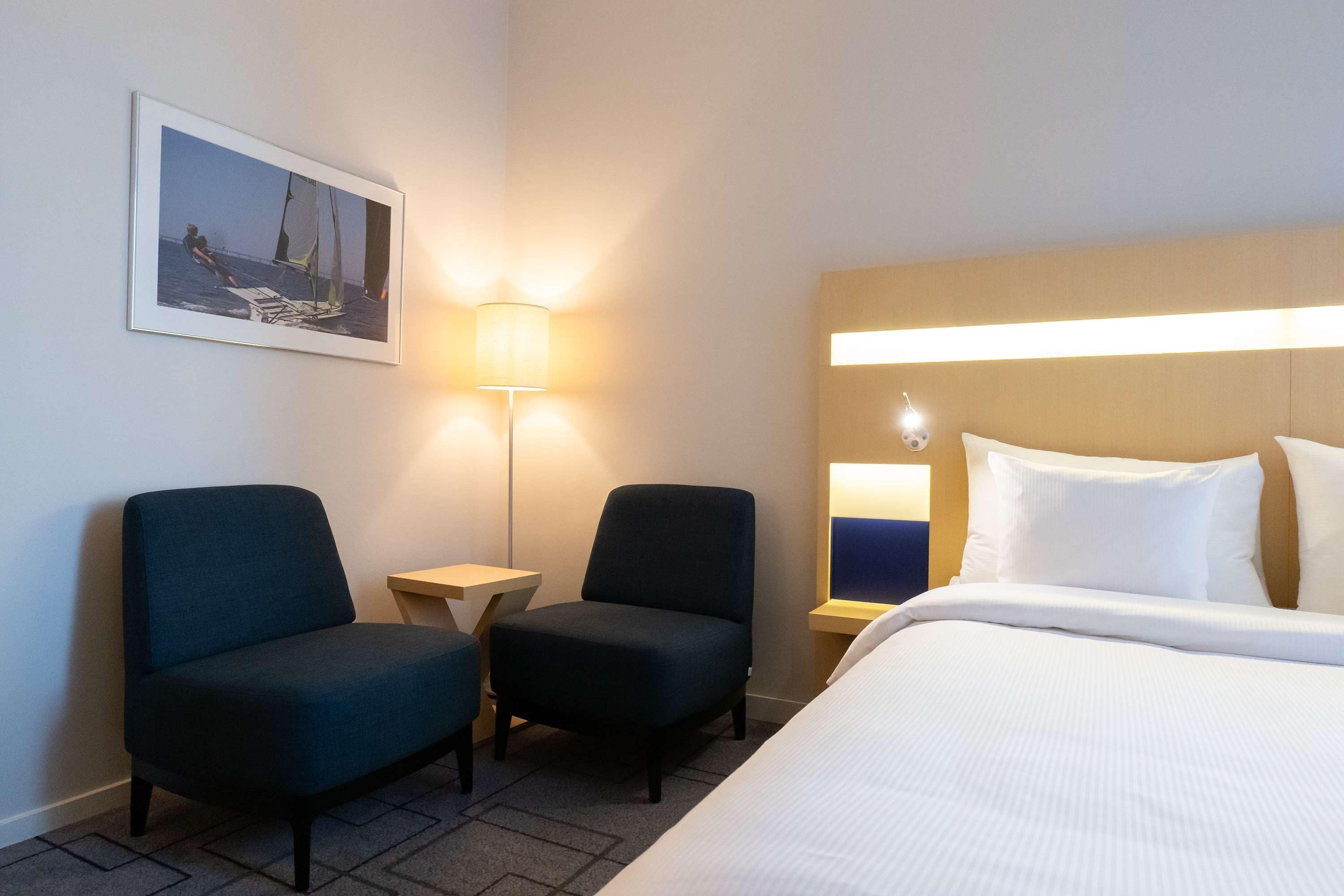 Quarto de hotel com uma cama, duas poltronas, uma mesinha, um abajur e um quadro na parede.