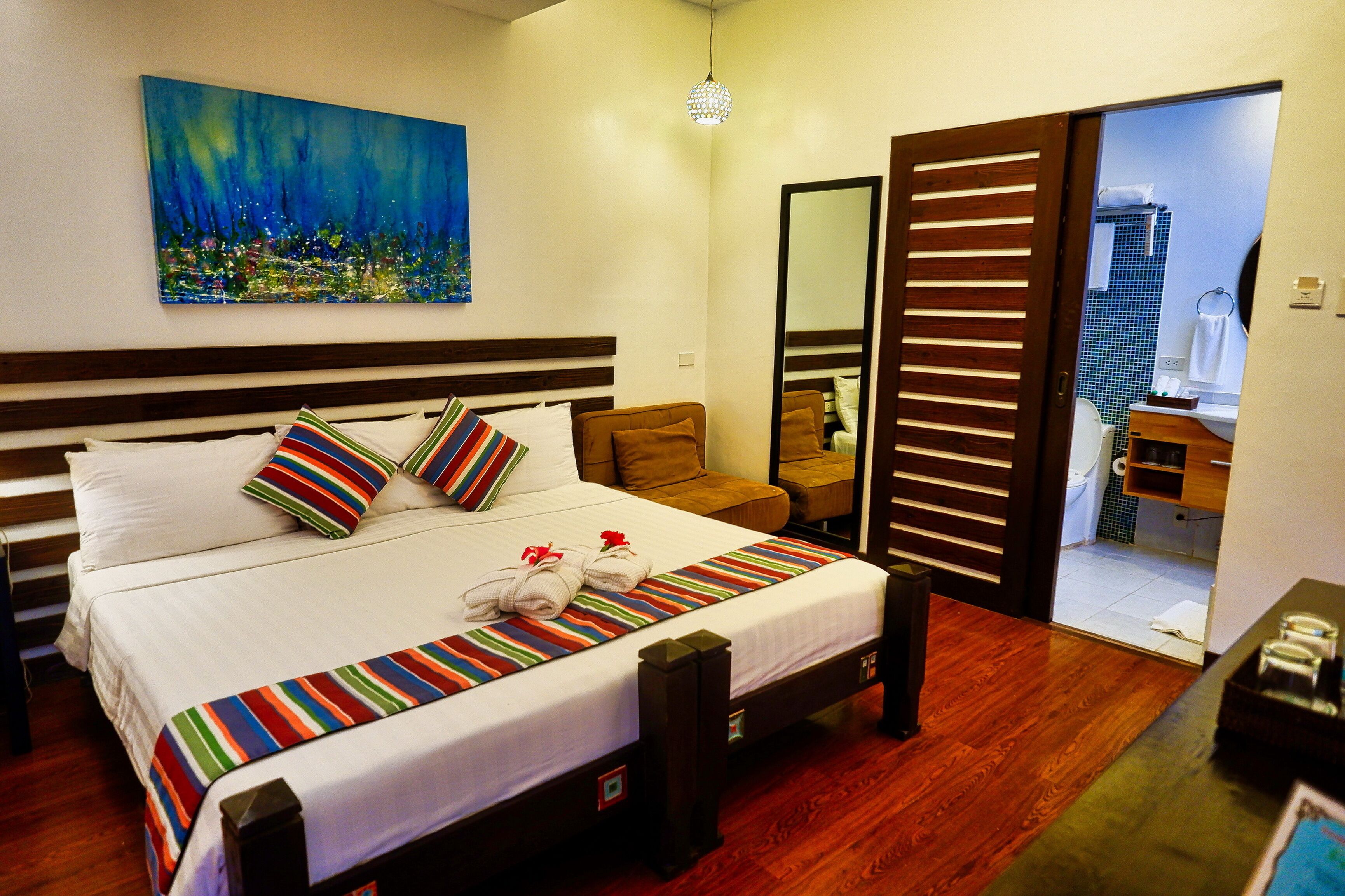 Quarto de hotel com uma cama grande, uma cadeira, banheiro visível por uma porta aberta e um quadro na parede.