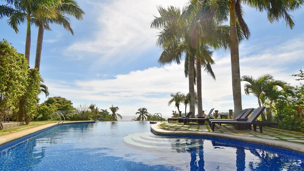 Piscina azul límpida cercada por palmeiras e espreguiçadeiras, com vista para o oceano ao fundo.