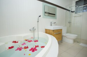 Uma casa de banho com uma banheira cheia de água e flores flutuantes, cabine de duche em vidro, sanita branca e toucador de madeira.