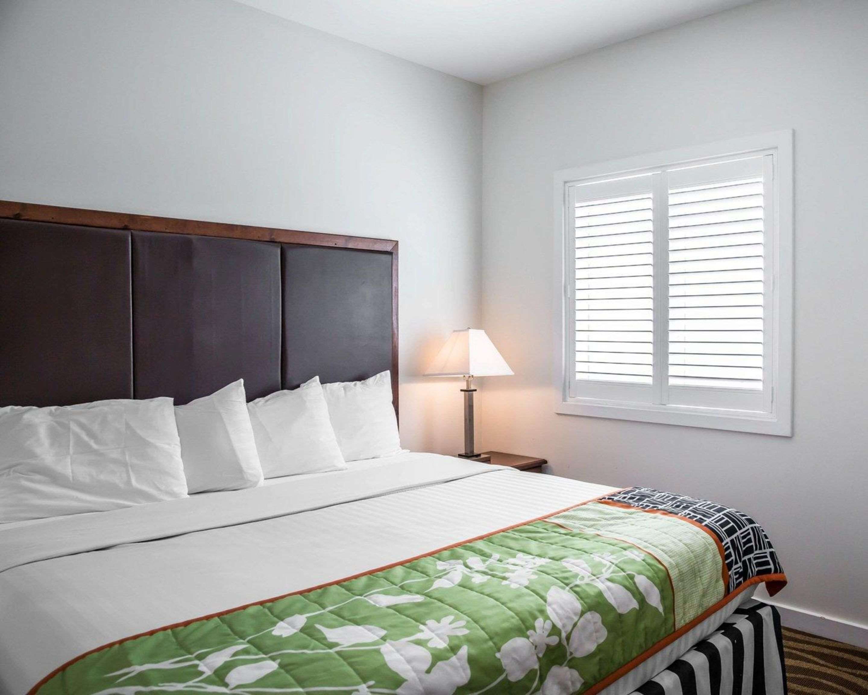 Uma cama bem arrumada com colcha floral verde e almofadas brancas, cabeceira de madeira, luminária de mesa e janela com persianas.