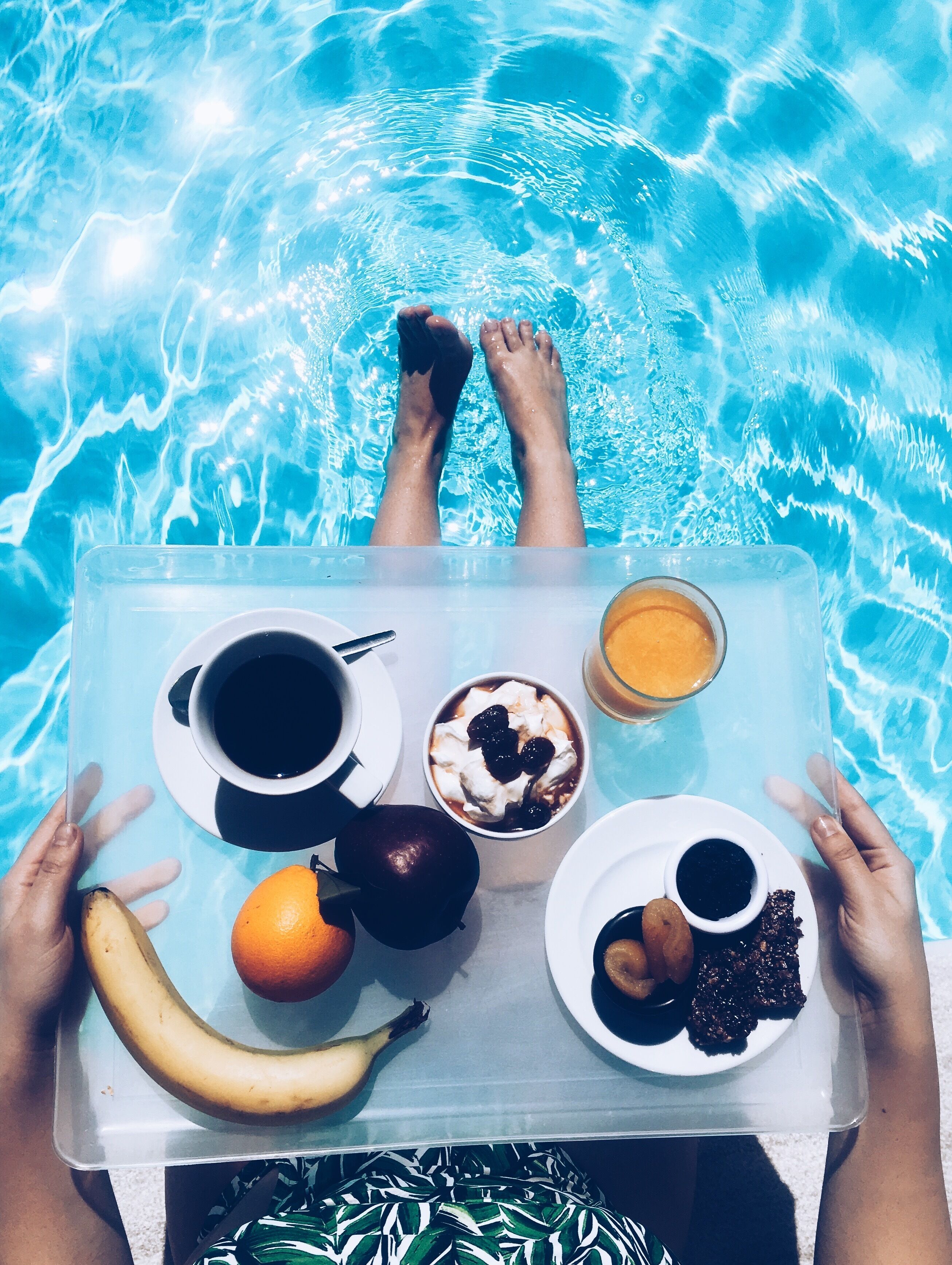 Vista da piscina com uma bandeja contendo comida e bebidas, incluindo uma banana, uma laranja e uma tigela de sobremesa.