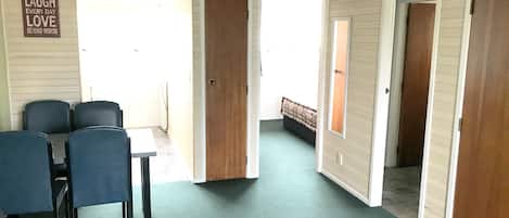Quarto com uma mesa de jantar e cadeiras, um sofá e uma pequena mesa com um controle remoto. Há duas portas que dão para outros cômodos.