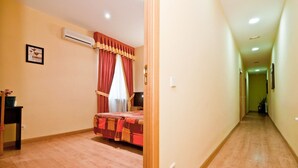 Quarto de hotel com cama, escrivaninha e ar condicionado. O quarto possui piso de madeira e um corredor que leva a outro quarto.