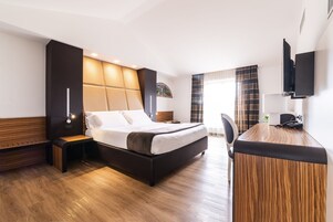 Quarto de hotel moderno com piso de madeira, cama com roupa de cama branca, escrivaninha com televisão de tela plana e uma grande janela com cortinas.