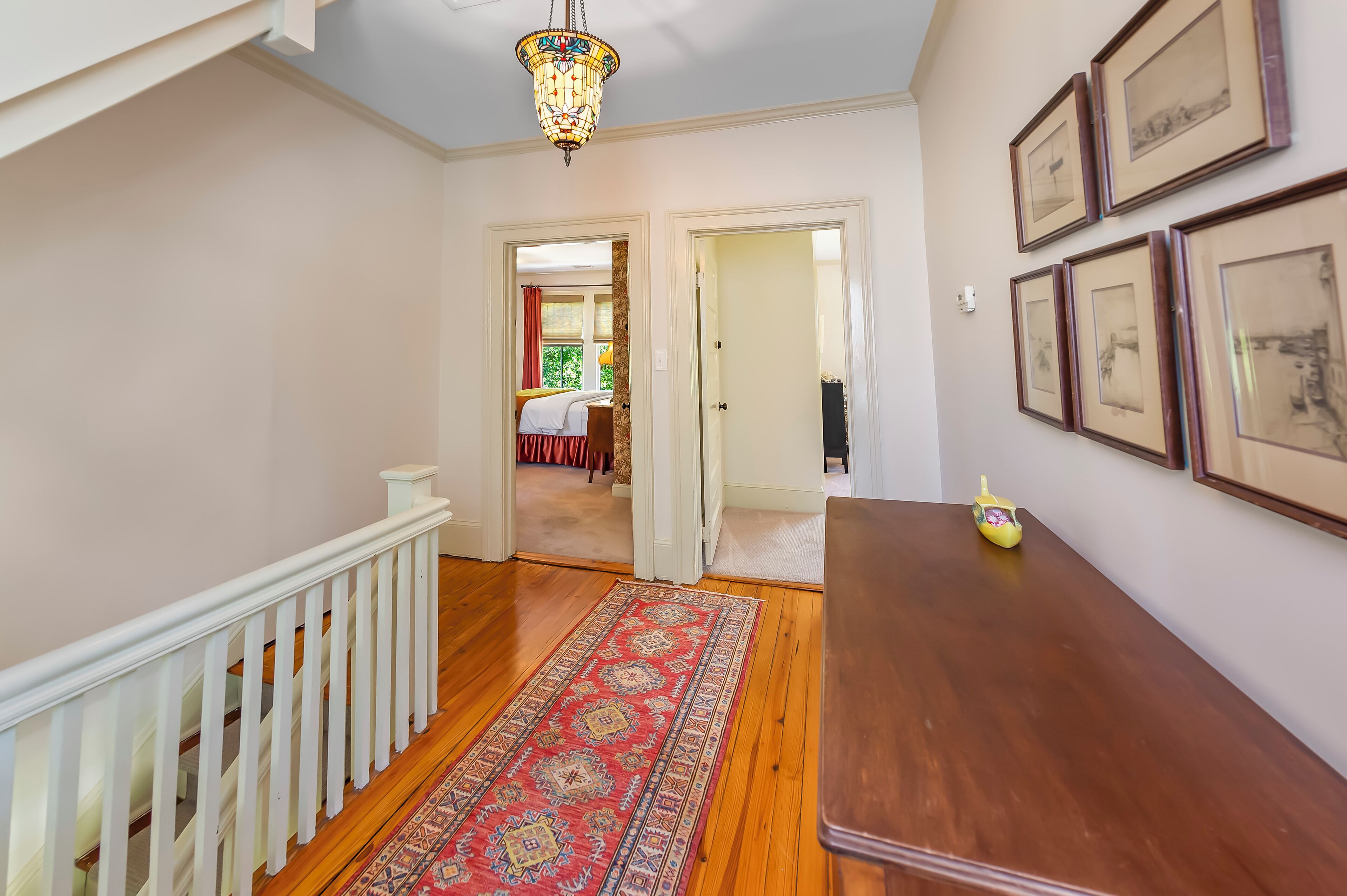 Um corredor com piso de madeira, tapete vermelho estampado, corrimão branco e um candelabro.