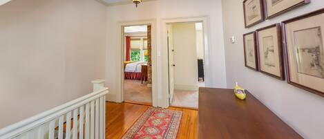 Um corredor com piso de madeira, tapete vermelho estampado, corrimão branco e um candelabro.
