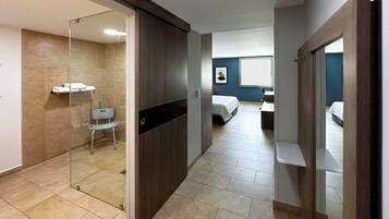 Quarto de hotel com chuveiro em cabine de vidro, uma cama, uma escrivaninha e uma cadeira.