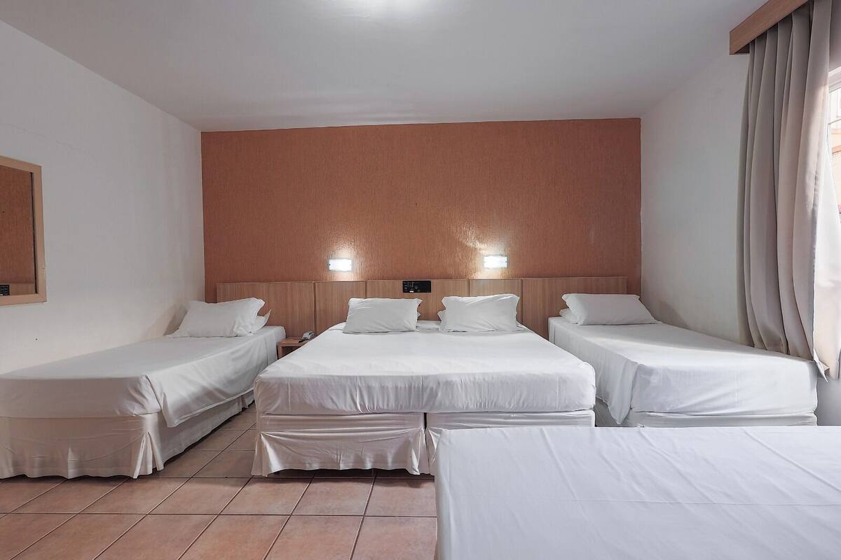 Quarto de hotel com três camas de solteiro, cabeceira de madeira, mesa de cabeceira pequena com abajur e um espelho grande na parede.