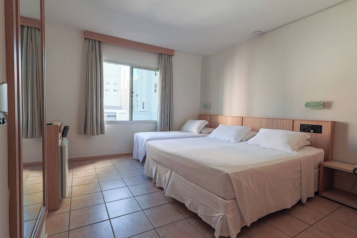 Quarto de hotel com duas camas, uma janela com cortinas, uma mesa de cabeceira e painel de controle fixado na parede.