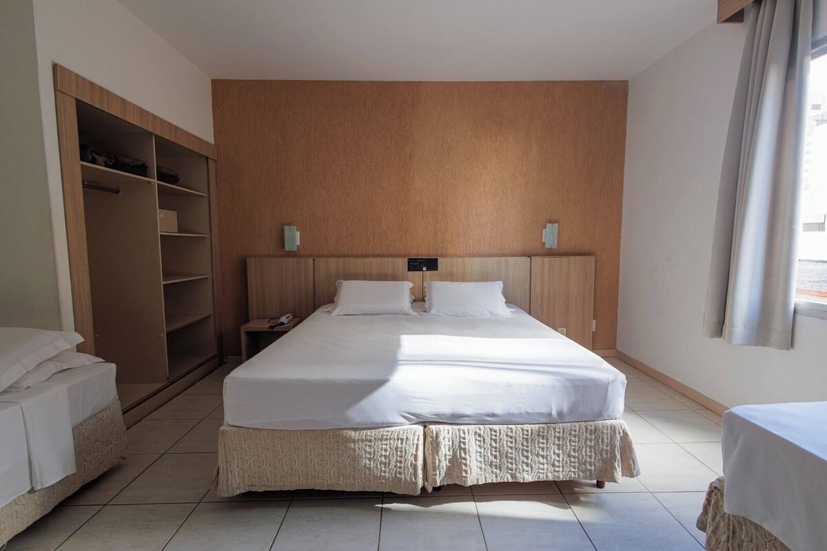 Quarto de hotel com cama grande, guarda-roupa, mesa de cabeceira com telefone e janela com cortinas.